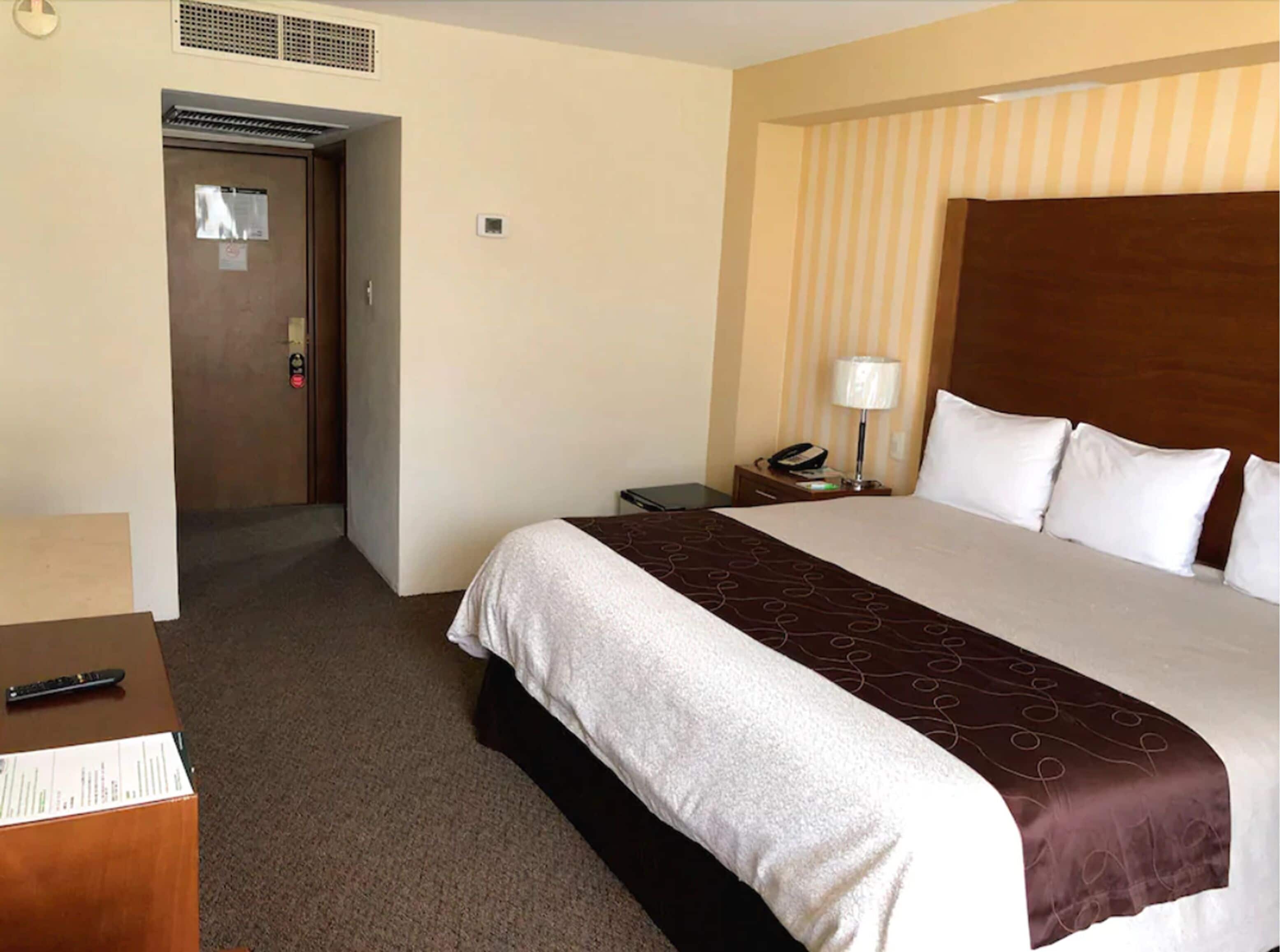 Quarto de hotel com uma cama grande, uma mesa de cabeceira, uma luminária e uma porta para um corredor.