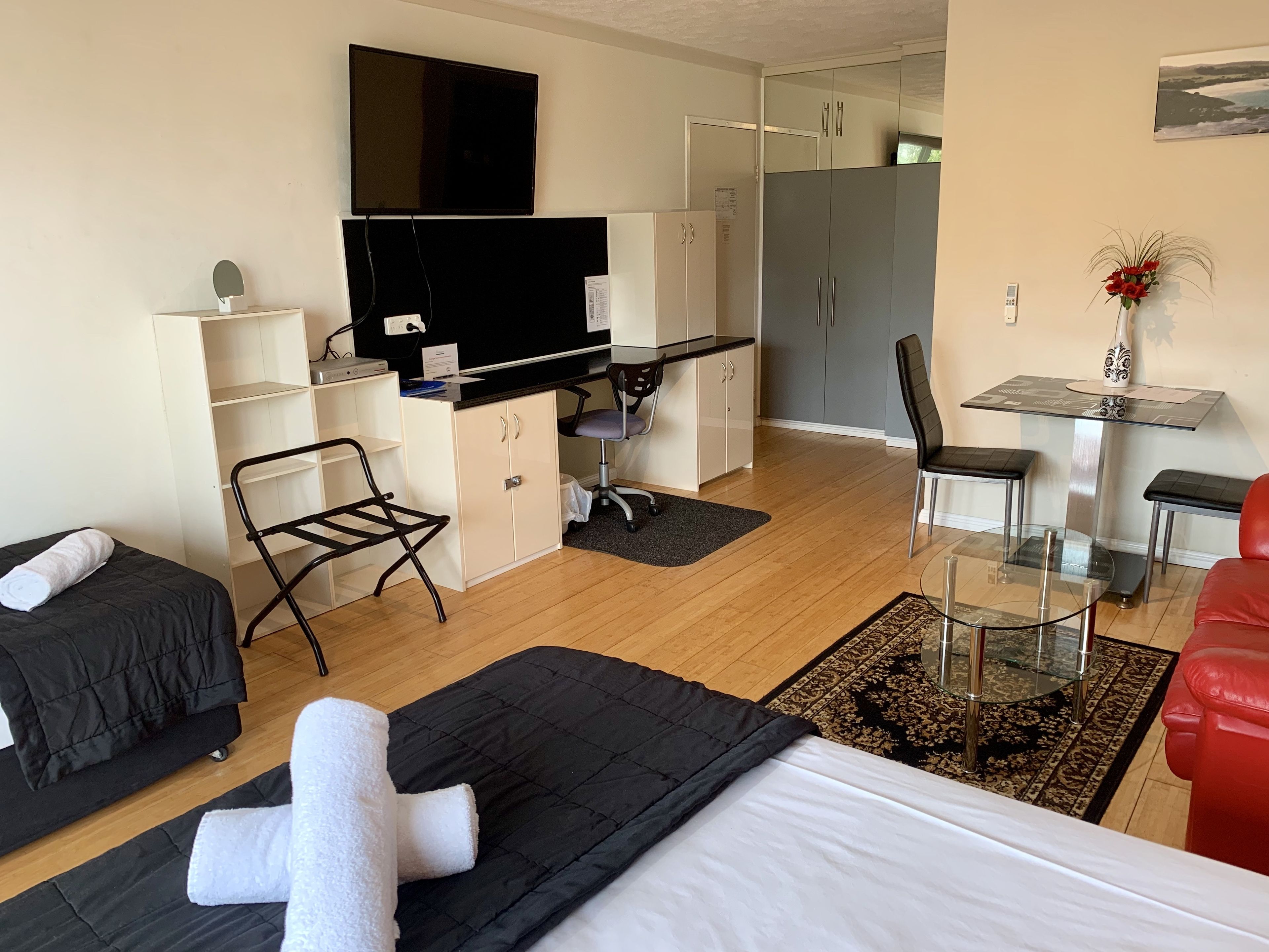 Quarto de hotel compacto com cama, escrivaninha, cadeira, televisão e pequena área de refeições.
