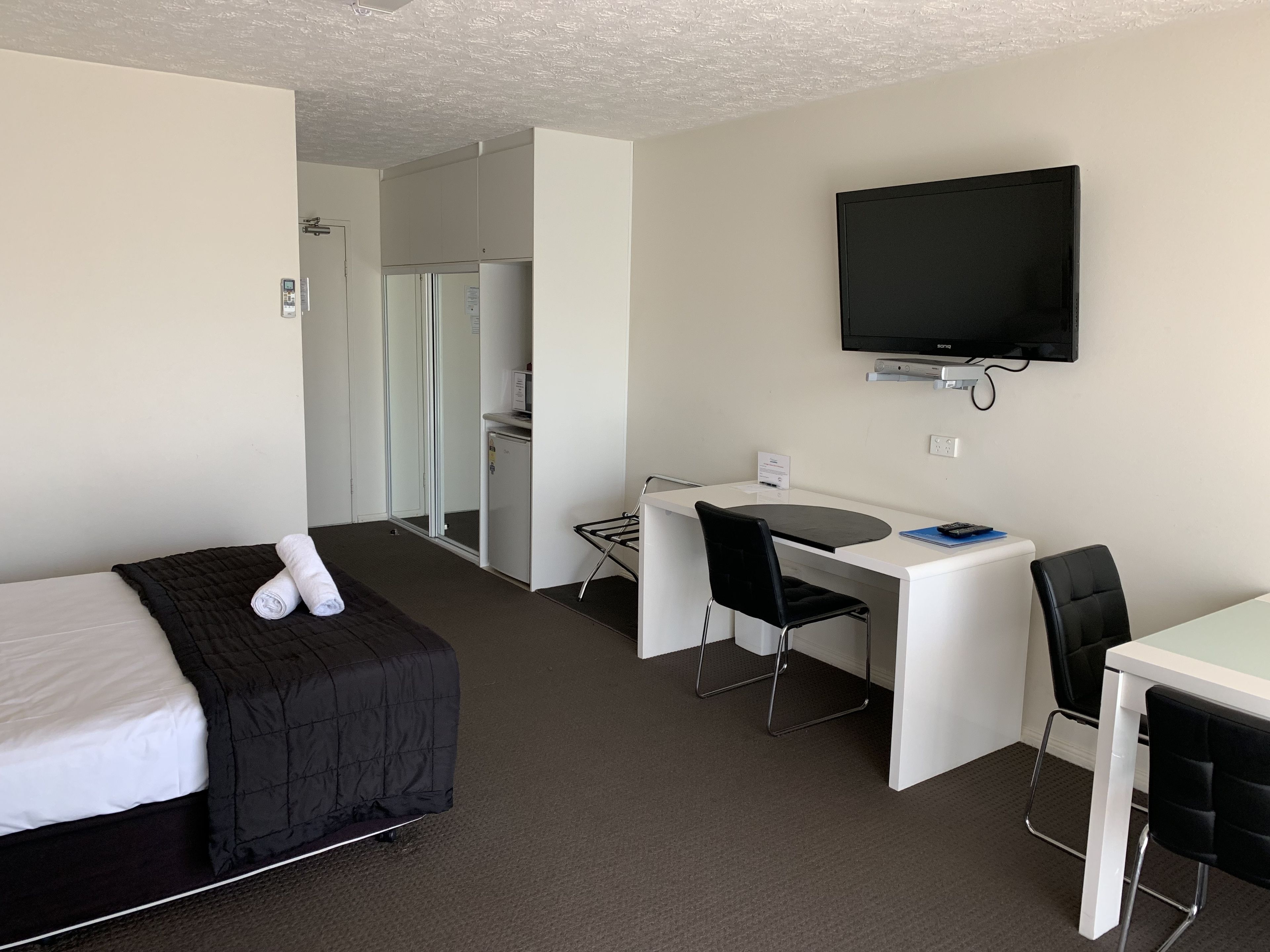 Quarto de hotel com cama, escrivaninha, cadeira, televisão e um pequeno frigorífico.