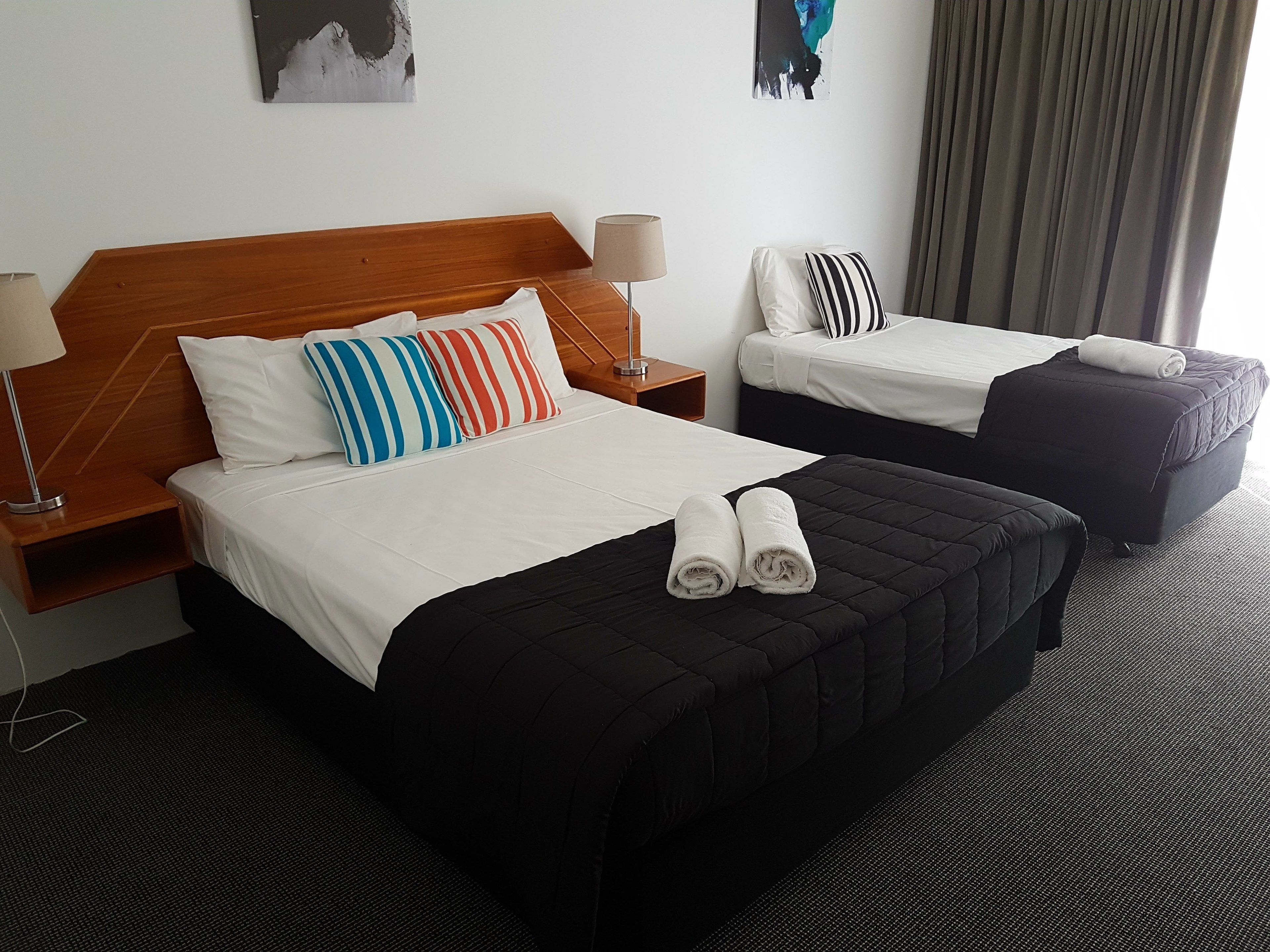 Quarto de hotel com uma cama grande, uma cama menor, duas mesas de cabeceira e uma janela com cortinas.