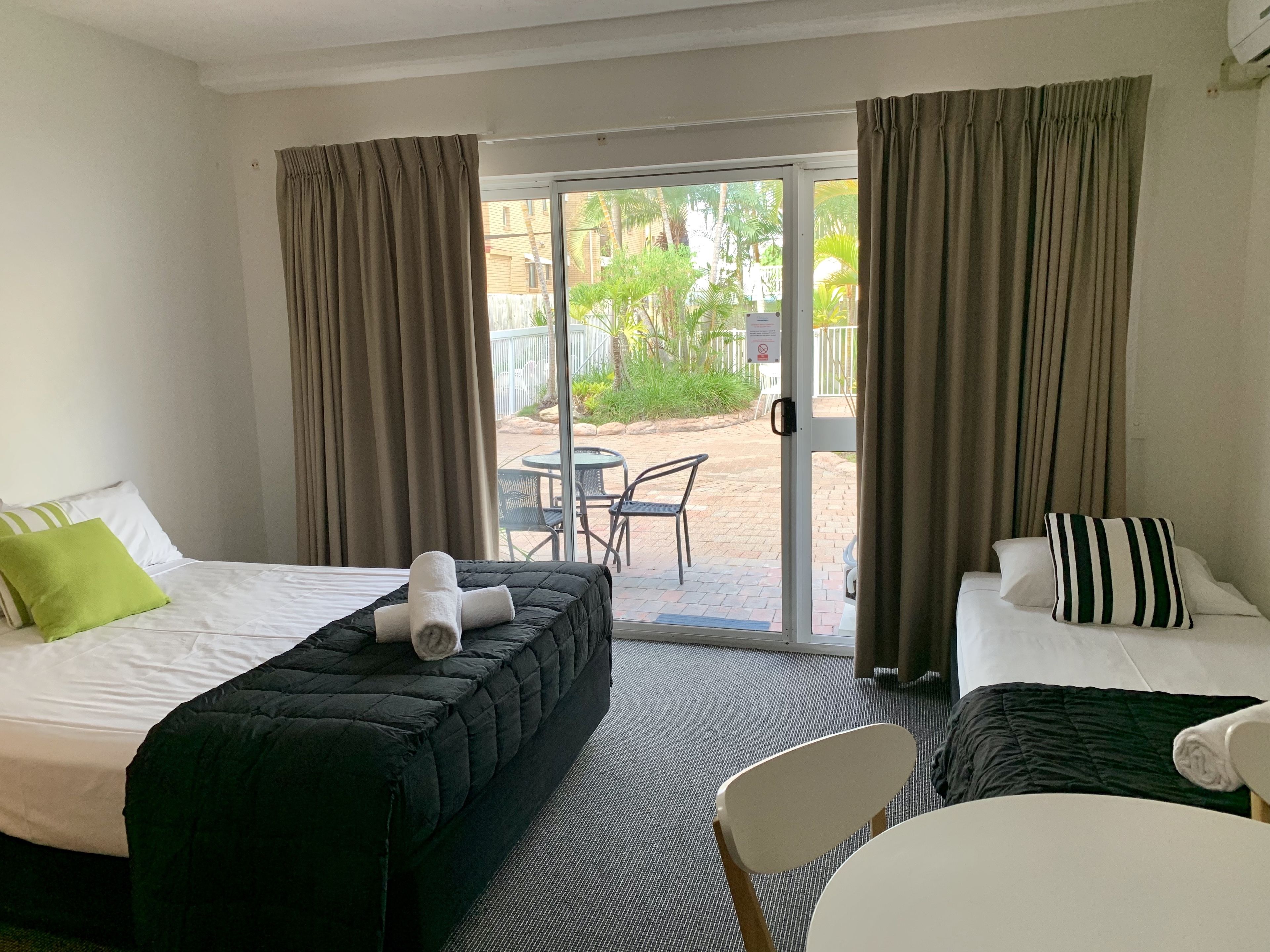 Quarto de hotel com duas camas, uma mesa pequena e uma porta de vidro deslizante para uma área de pátio ao ar livre.