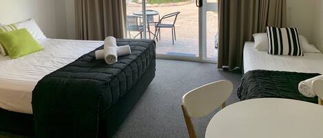 Quarto de hotel com duas camas, uma mesa pequena e uma porta de vidro deslizante para uma área de pátio ao ar livre.