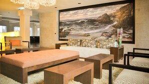 Quarto de hotel moderno com uma grande pintura vista para o oceano, um sofá, um banco de madeira e uma área de jantar com mesa e cadeiras.