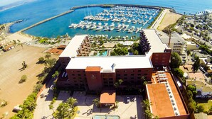 Vista aérea de um hotel com marina, destacando um edifício grande com telhado plano, piscina e área de estacionamento.