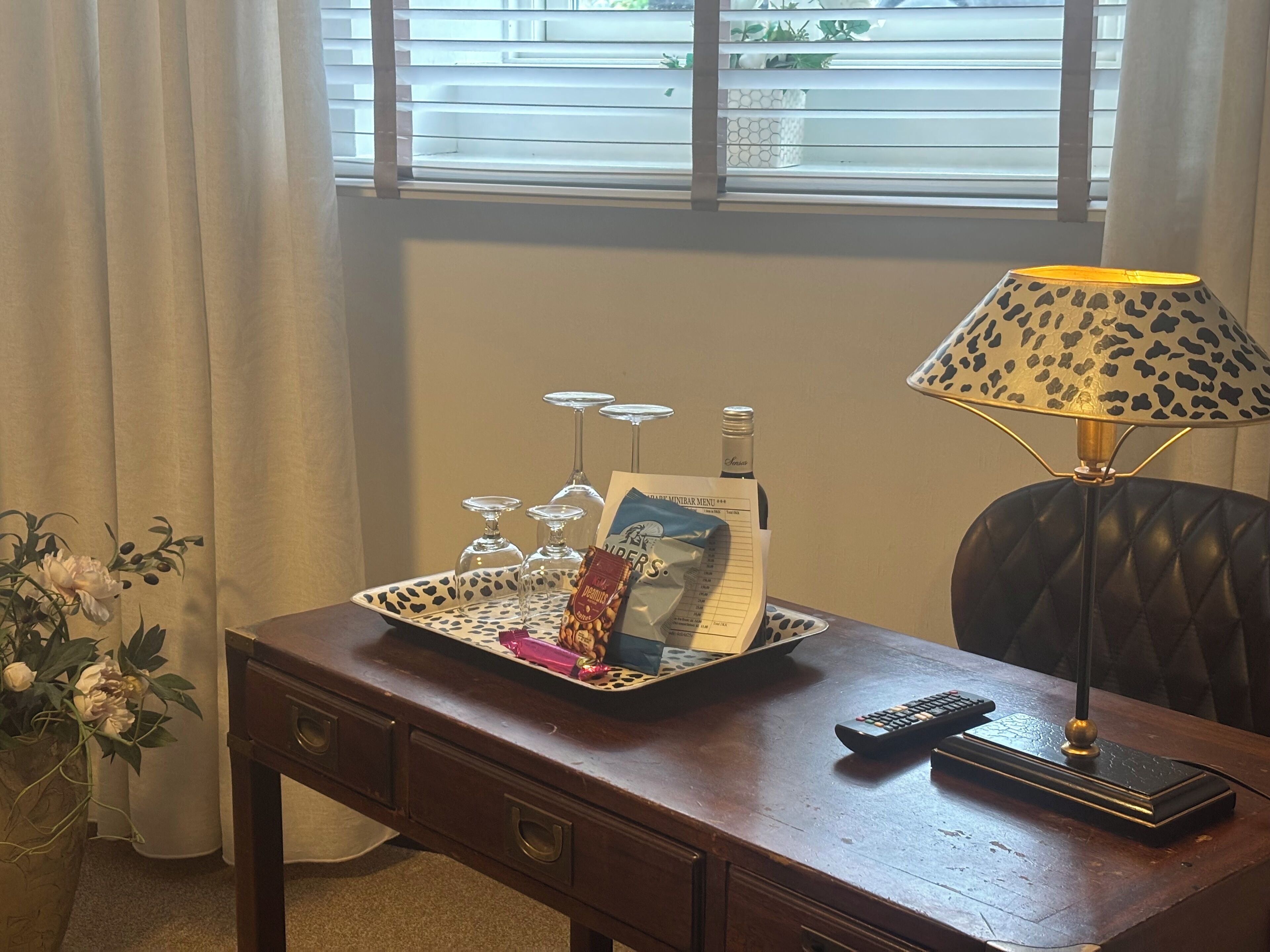 Uma escrivaninha de madeira com um suporte contendo copos de vinho, uma garrafa e um cardápio. Uma luminária com estampa de leopardo e um controle remoto estão sobre a escrivaninha.