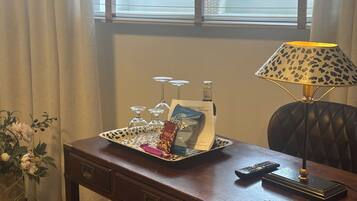 Uma escrivaninha de madeira com um suporte contendo copos de vinho, uma garrafa e um cardápio. Uma luminária com estampa de leopardo e um controle remoto estão sobre a escrivaninha.