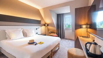 Quarto de hotel moderno com uma cama grande, uma mesa com chaleira, uma cadeira e vista para o exterior.