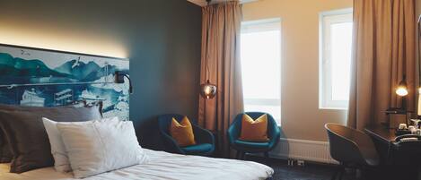 Quarto de hotel com cama, duas cadeiras, escrivaninha e janela com cortinas.