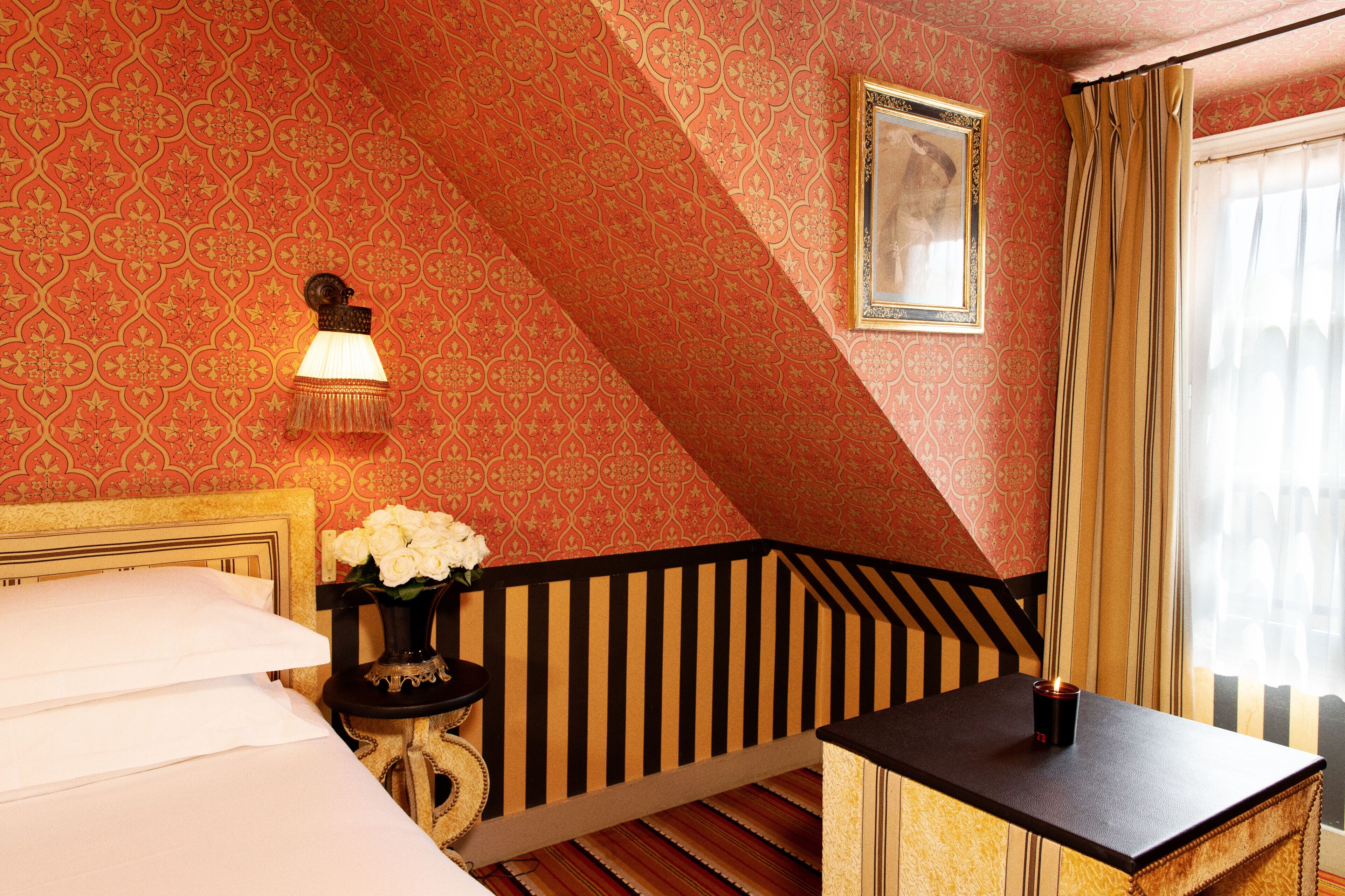 Quarto de hotel com parede vermelha estampada, cama com roupa de cama branca, mesa de cabeceira com abajur e flores, e vista para o exterior através de uma janela com cortinas.