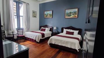 Quarto de hotel com duas camas, um ventilador de teto, uma mesa e uma janela com cortinas.
