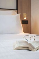Cama arrumada com travesseiro branco e um livro aberto com óculos em cima.