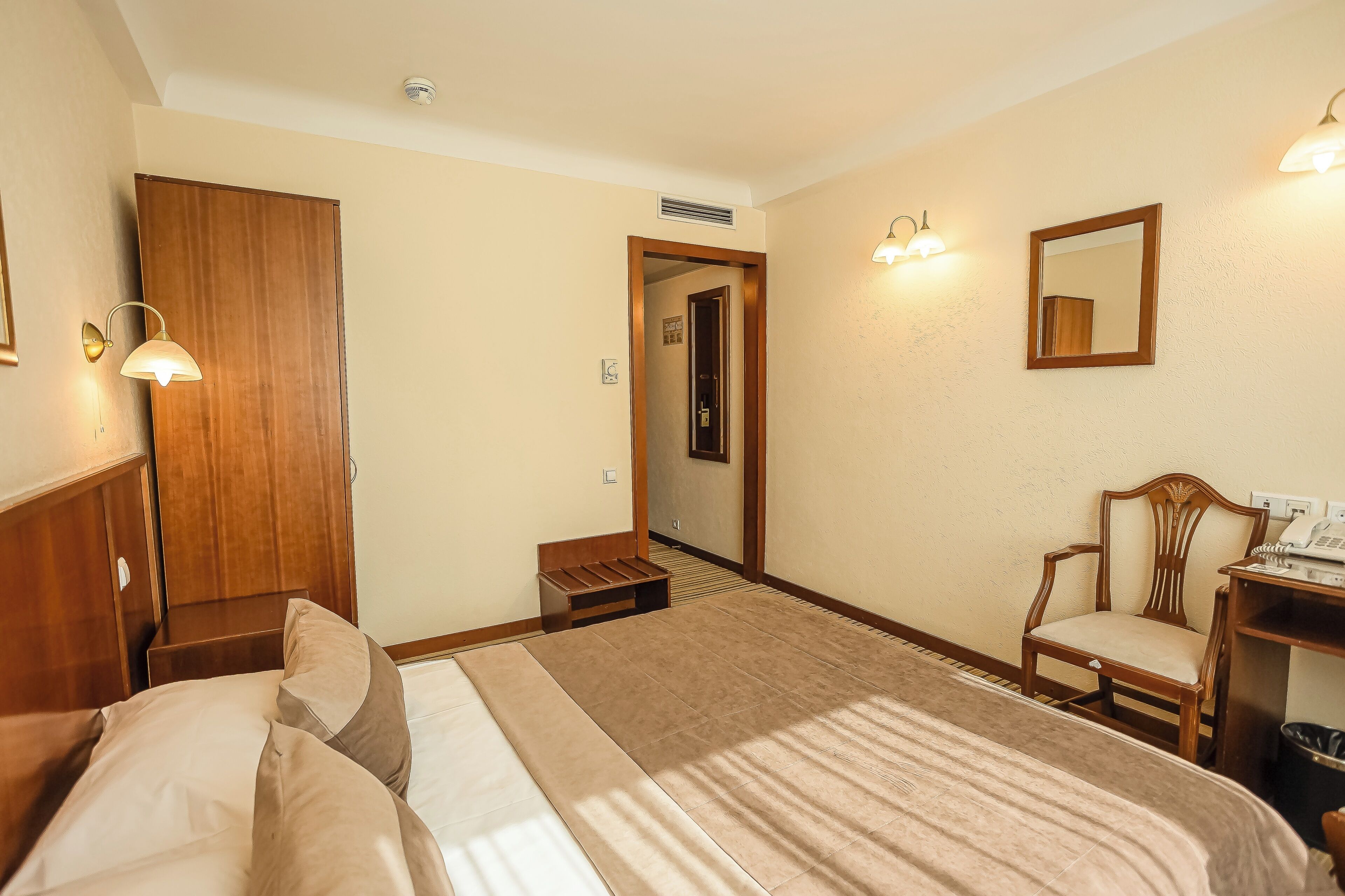 Quarto de hotel com cama, escrivaninha com telefone, cadeira, espelho e porta para outro cômodo.