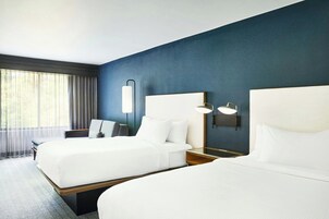 Quarto de hotel com duas camas, uma janela grande com cortinas, uma mesa de cabeceira com abajur e luminária na parede.