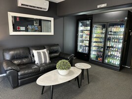 Uma área de estar moderna com um sofá de couro preto, uma mesa redonda pequena com uma planta em vaso e uma geladeira repleta de bebidas.