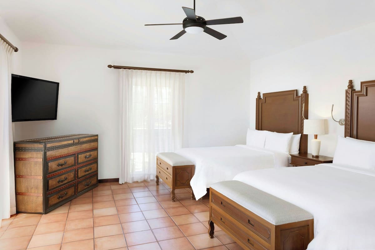 Quarto de hotel com duas camas, uma cômoda de madeira, um ventilador de teto, televisão e uma janela com cortinas.