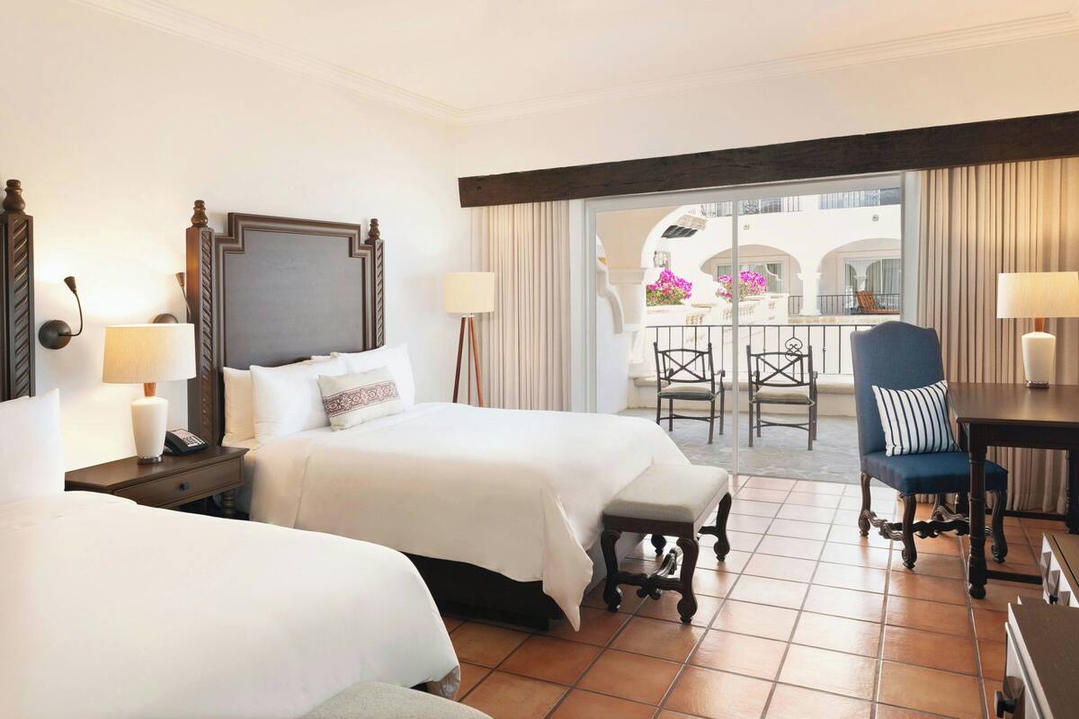 Quarto de hotel com duas camas, uma escrivaninha, uma cadeira e uma varanda com vista para a arquitetura do edifício.