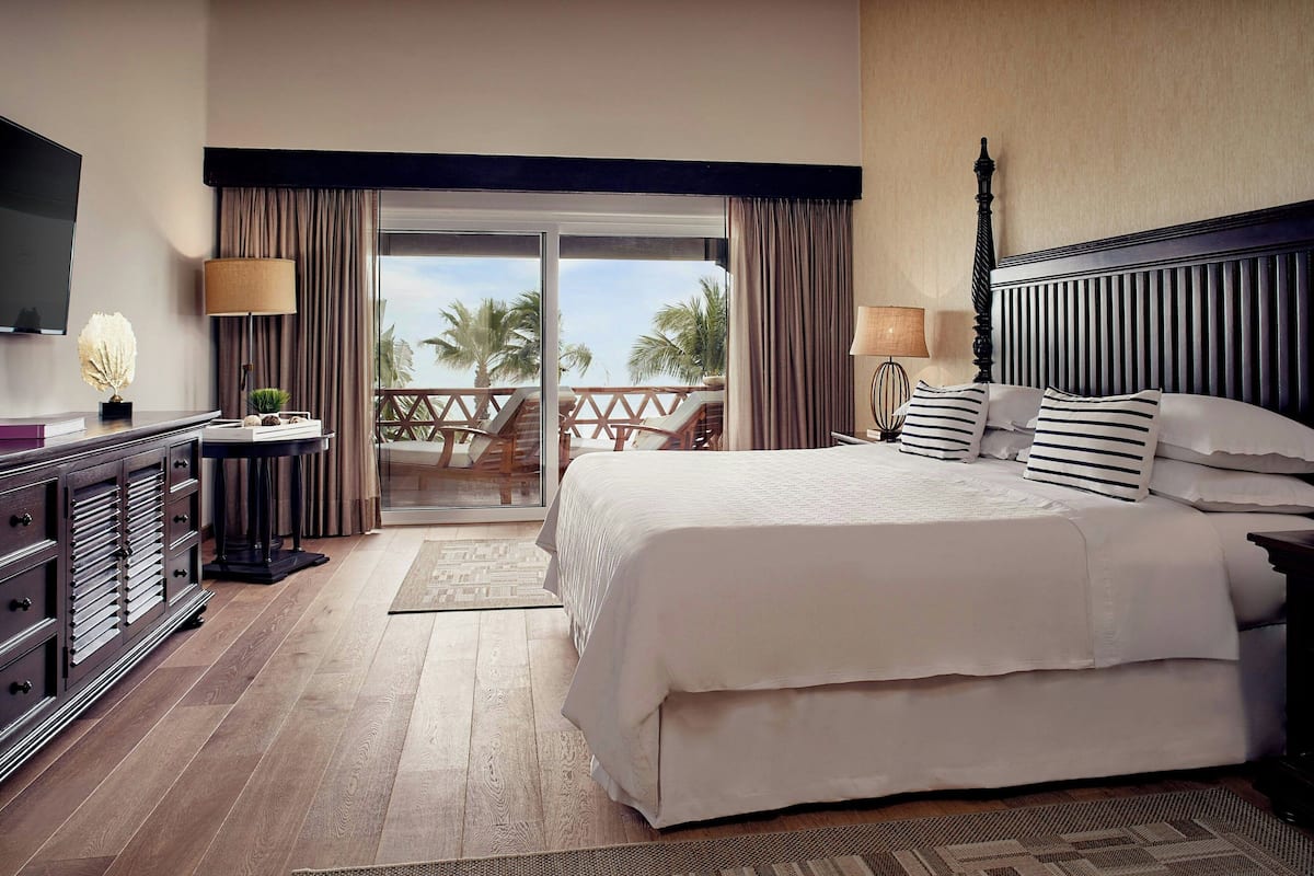 Quarto de hotel com uma cama grande, televisão, escrivaninha e varanda com vista para palmeiras.