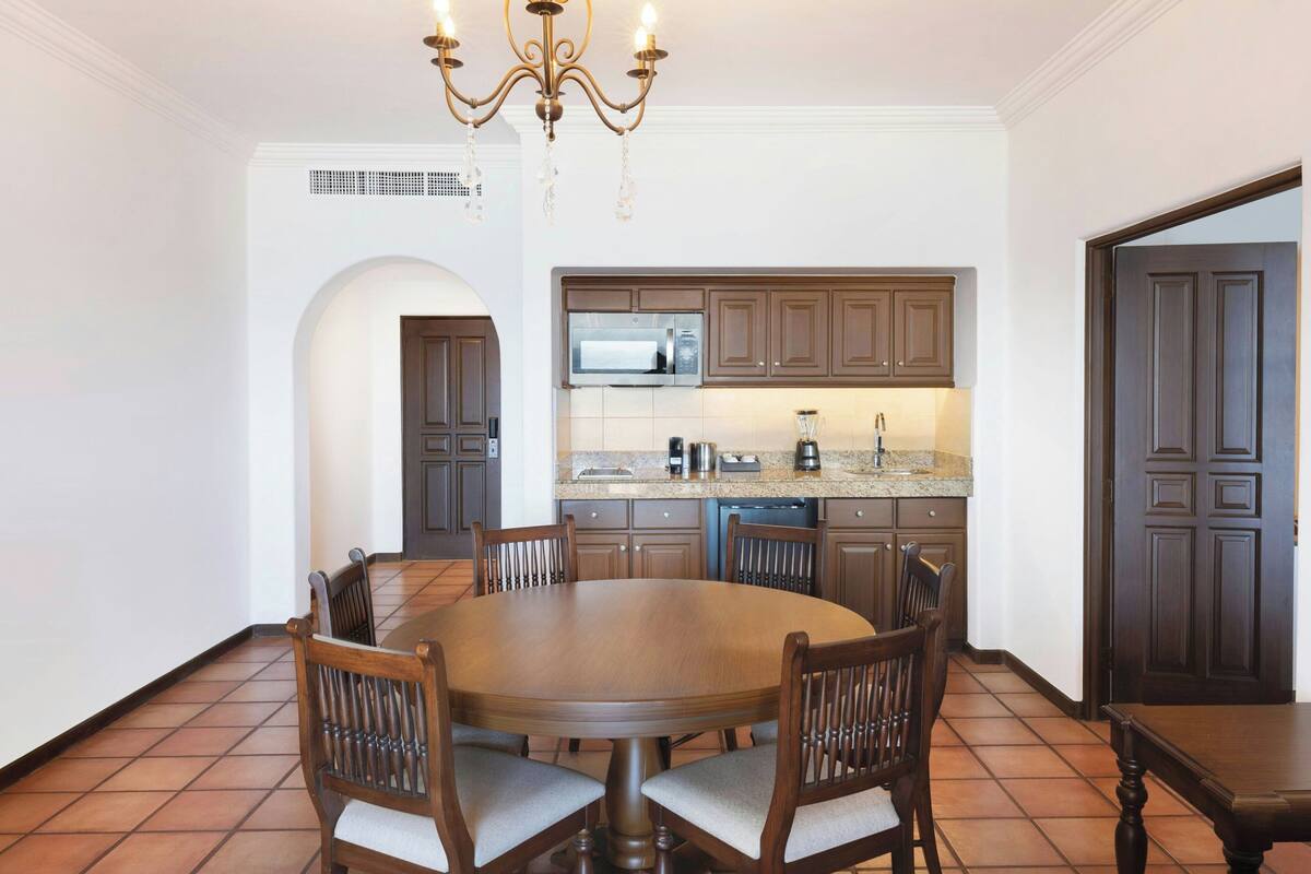 Uma sala de jantar com mesa redonda e cadeiras, uma cozinha com armários escuros e um micro-ondas, e um lustre.