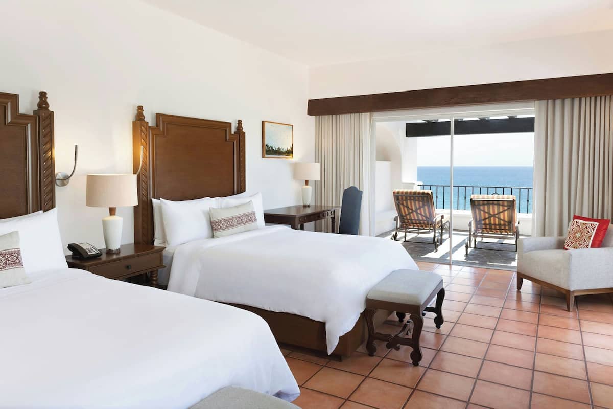 Quarto de hotel com duas camas, varanda com cadeiras e vista para o mar.
