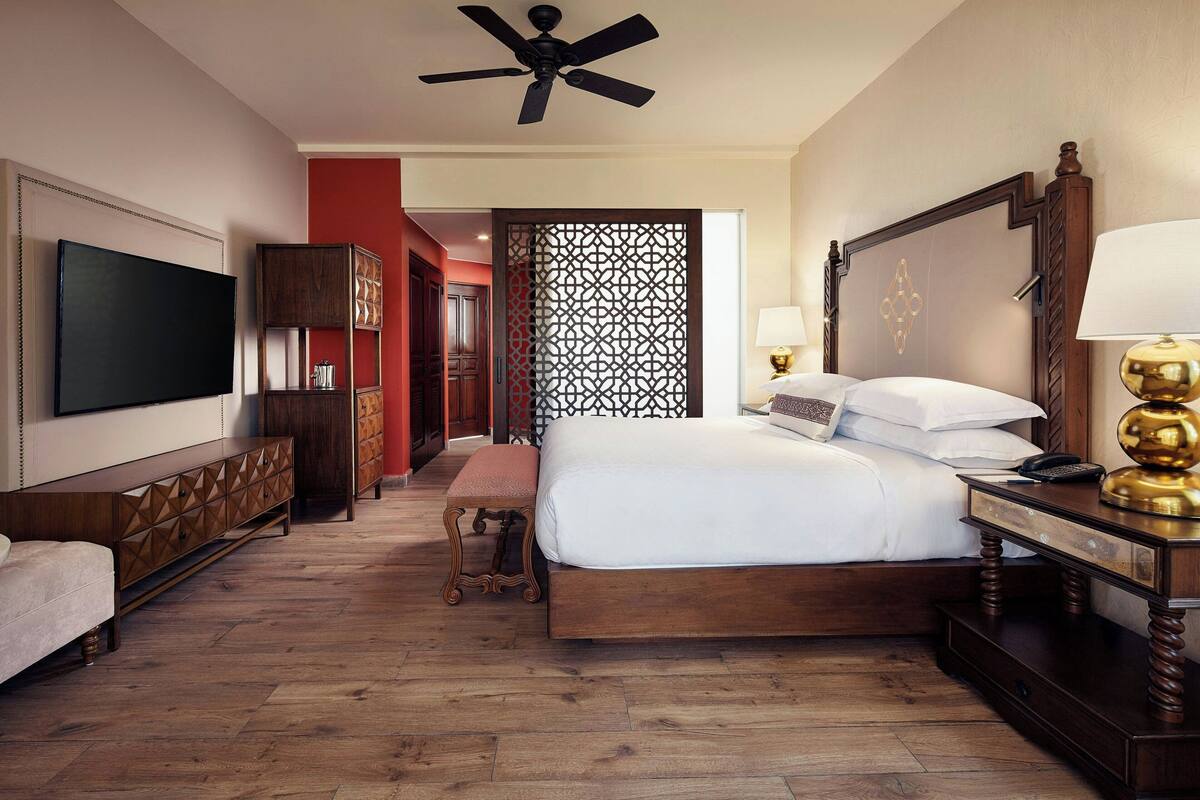 Quarto de hotel com cama, mesa de cabeceira, luminária, TV e piso de madeira.