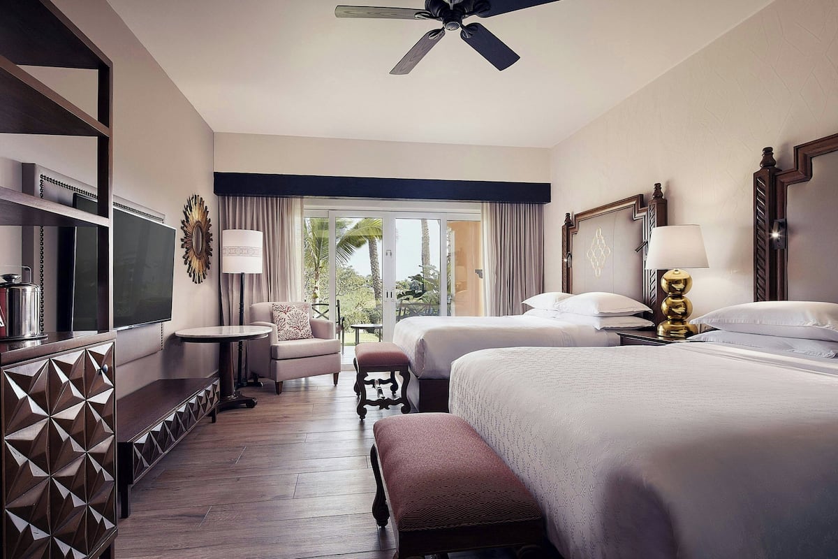 Quarto de hotel com duas camas, televisão, ventoinha no teto, uma mesinha e vista para o exterior.