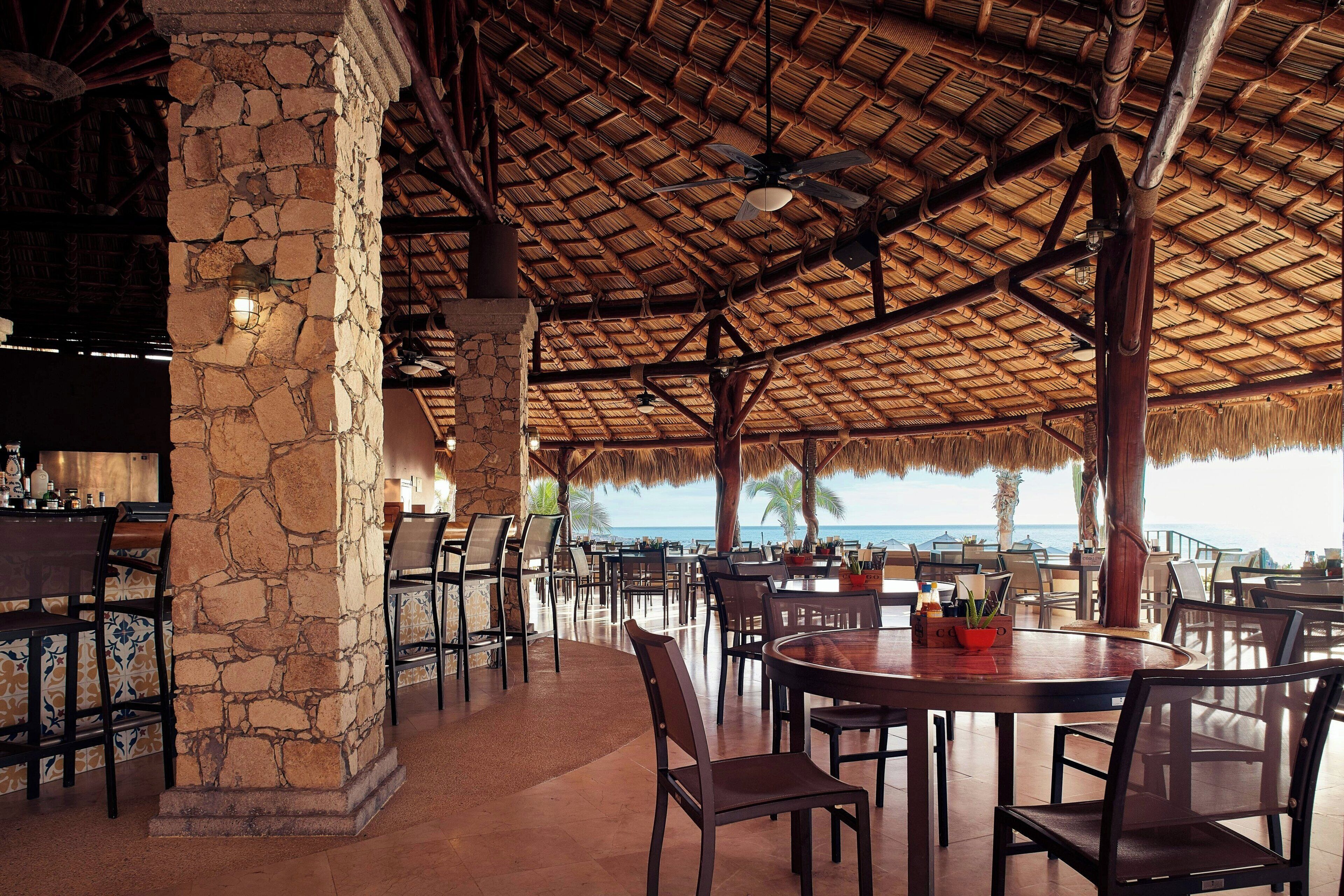 Um restaurante com telhado de palha, pilares de pedra e vista para o mar.