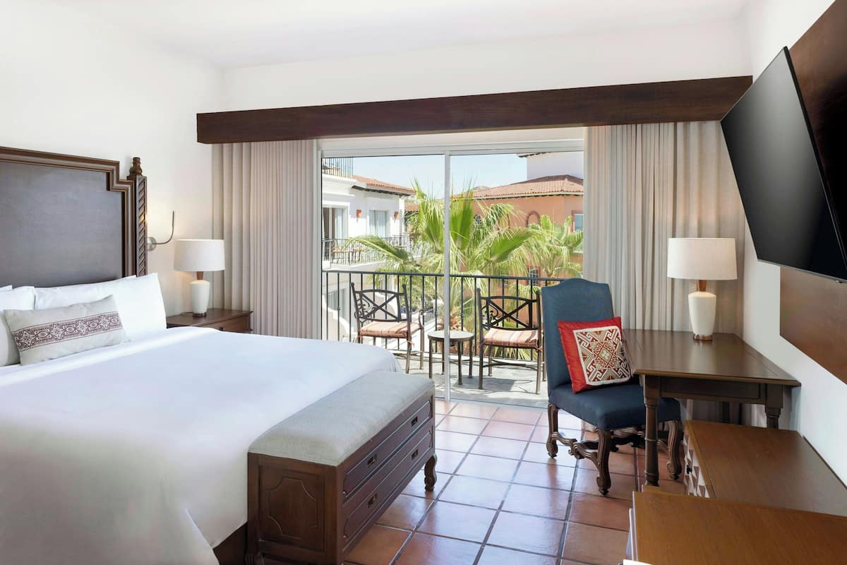 Quarto de hotel com uma cama grande, uma mesa de trabalho, uma cadeira, televisão e varanda com vista para palmeiras e edifícios.