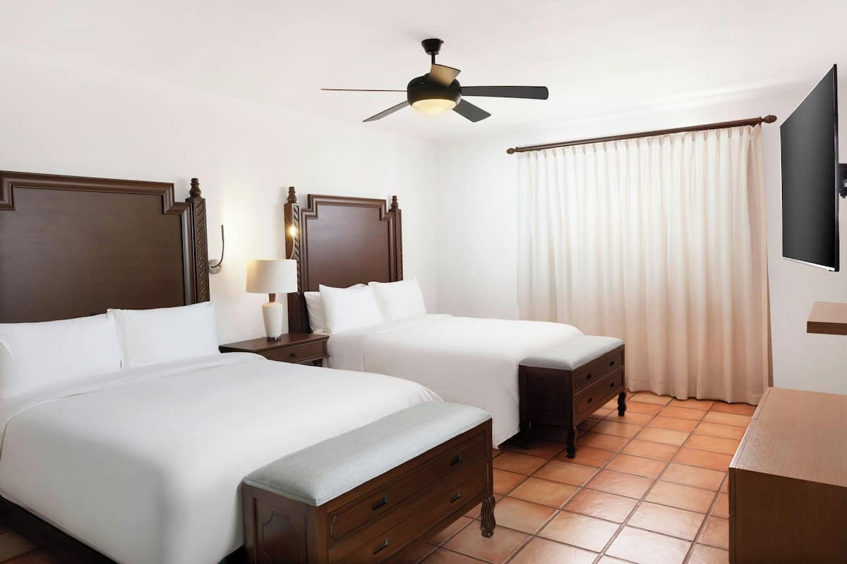Quarto de hotel com duas camas, um ventilador de teto, televisão e janela com cortinas.