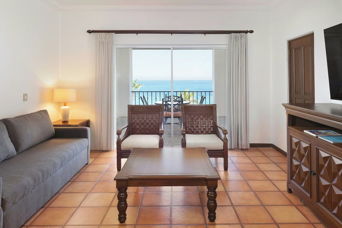 Quarto com sofá, mesa, duas cadeiras, televisão e vista para o mar por uma porta de correr.