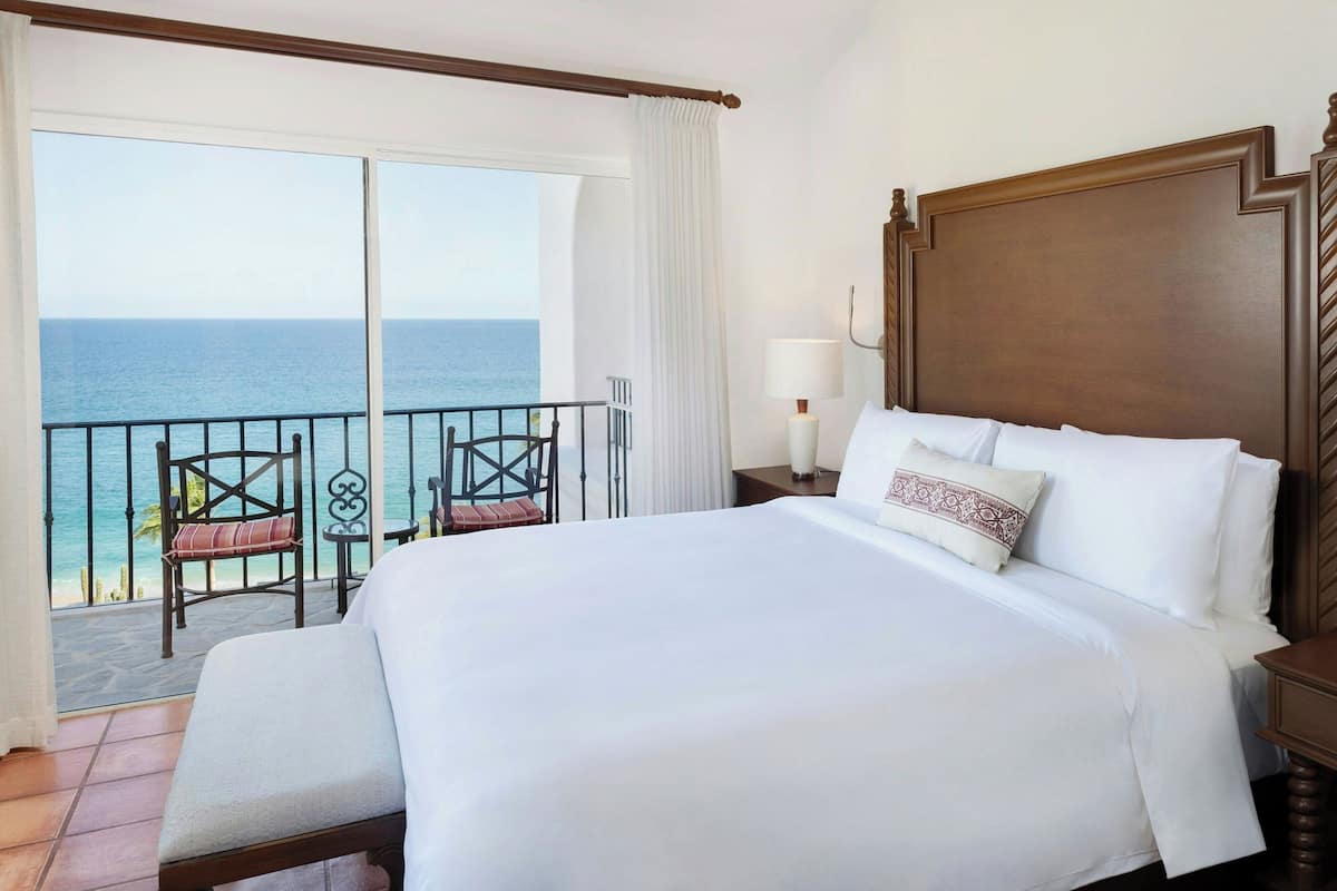 Quarto de hotel com uma cama grande, mesa de cabeceira, luminária e vista para o oceano.