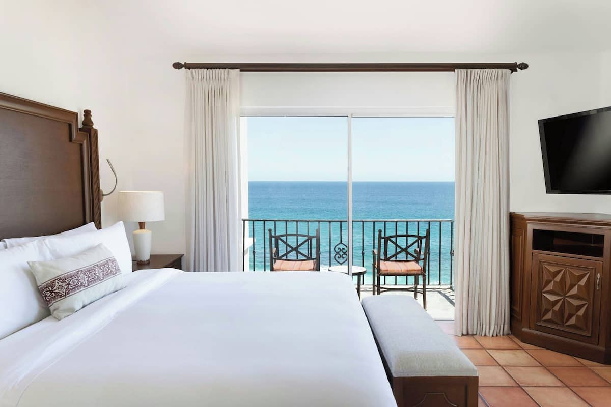 Quarto de hotel com uma cama grande, televisão, varanda vista para o mar e uma pequena mesa com cadeiras.