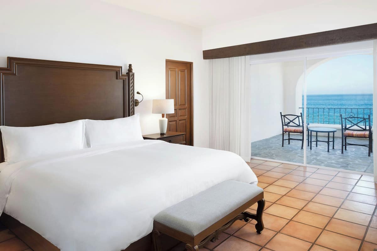 Quarto de hotel com cama grande, um banco, mesa de cabeceira com luminária e vista para o oceano por uma porta de correr.
