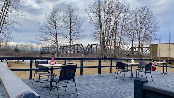 Um terraço ao ar livre com mesas e cadeiras, com vista para uma ponte e árvores.