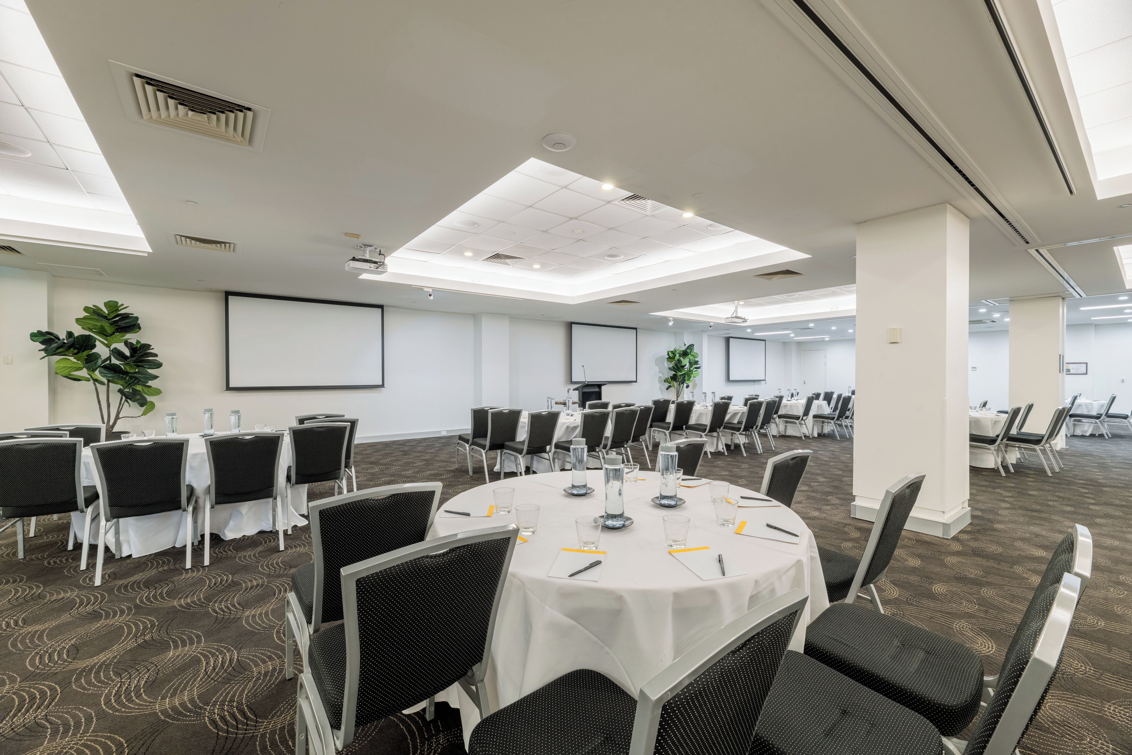 Sala de conferências com mesas redondas, cadeiras e tela de projeção.