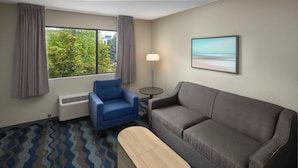 Quarto de hotel com uma janela grande, uma poltrona azul, um sofá cinzento, uma mesa redonda pequena e uma mesinha de cabeceira de madeira.
