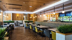 Restaurante moderno com piso de madeira, cadeiras amarelas e cinzas, e área de bar com revestimento de azulejos.