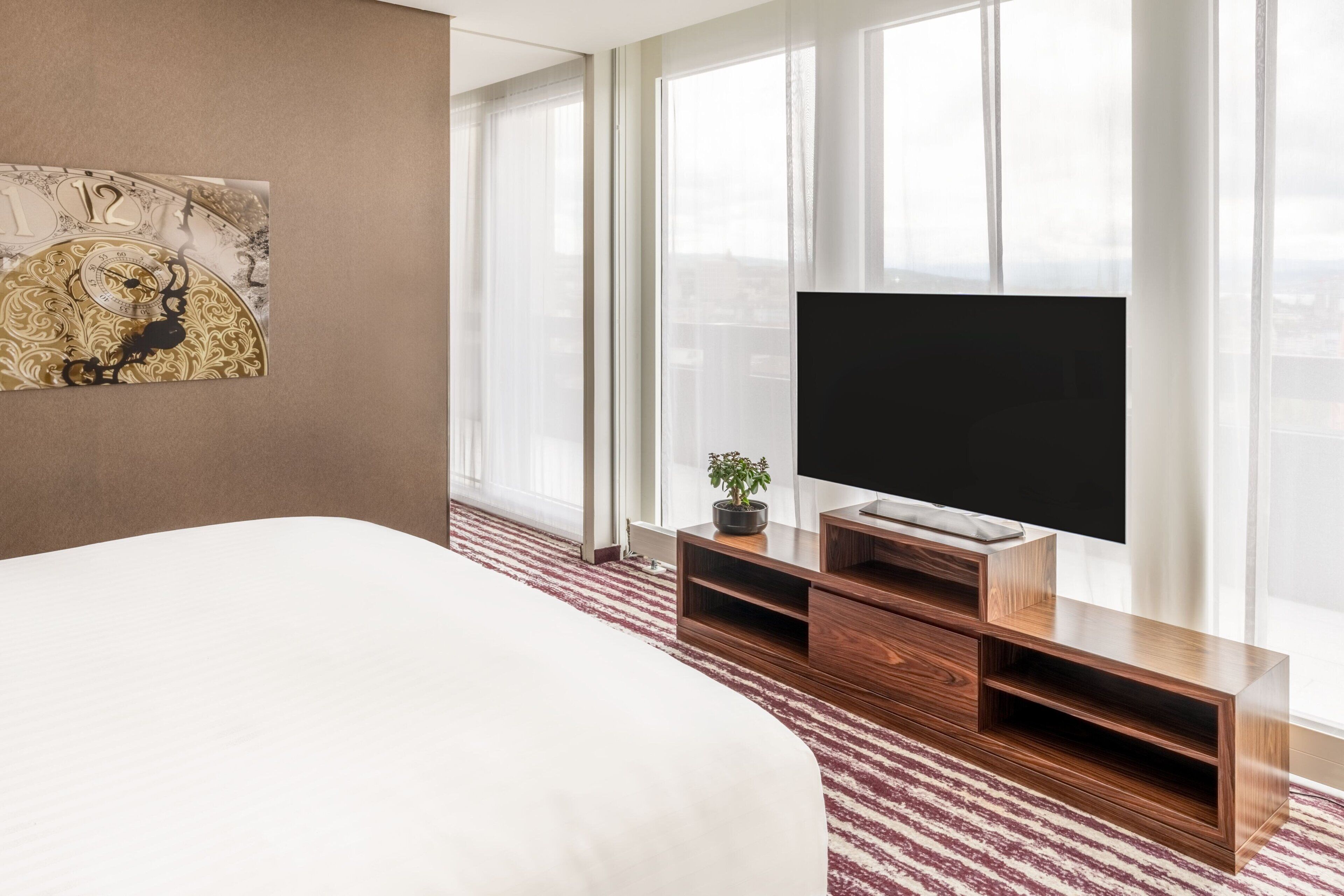 Quarto de hotel moderno com uma cama grande, uma TV de tela plana em um suporte de madeira e vista para a cidade através de grandes janelas.