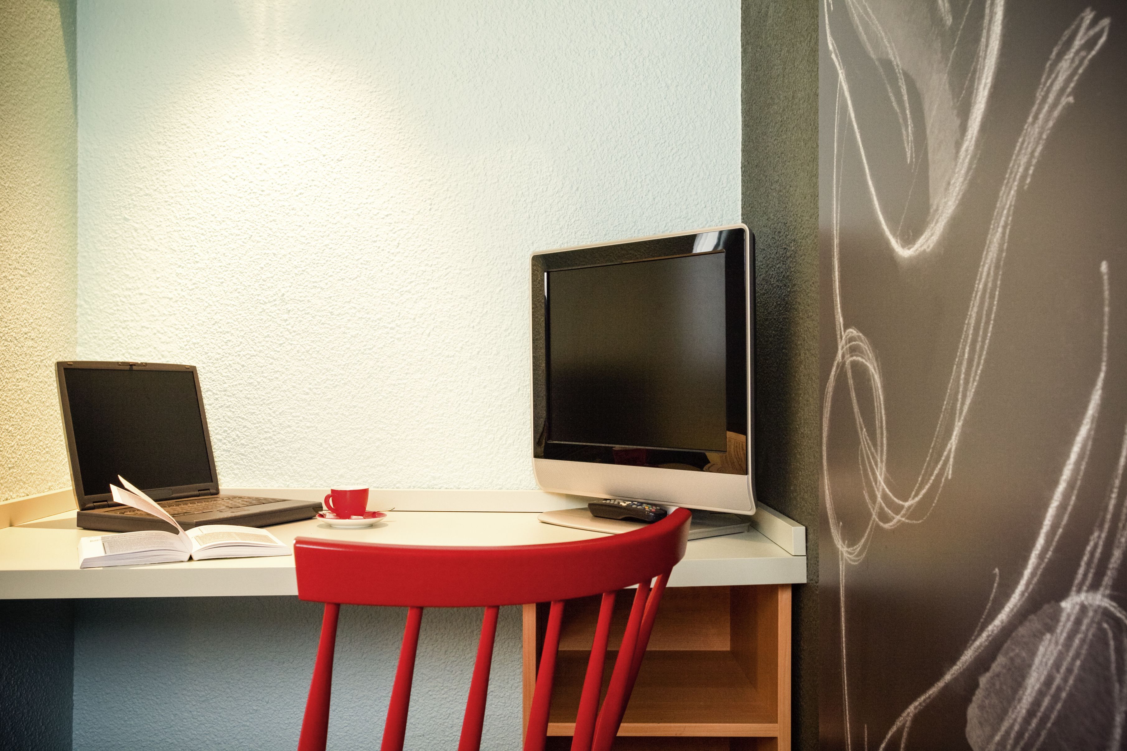Um espaço de trabalho com um laptop, um monitor de computador, uma cadeira vermelha, um livro e uma chávena sobre uma secretária branca, com uma parede texturizada ao fundo.