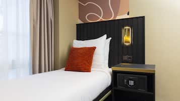 Quarto de hotel com cama, mesa de cabeceira com cofre, luminária na parede e janela com cortinas.