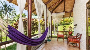 Uma área externa coberta com uma rede, cadeiras de madeira e vista para um jardim.
