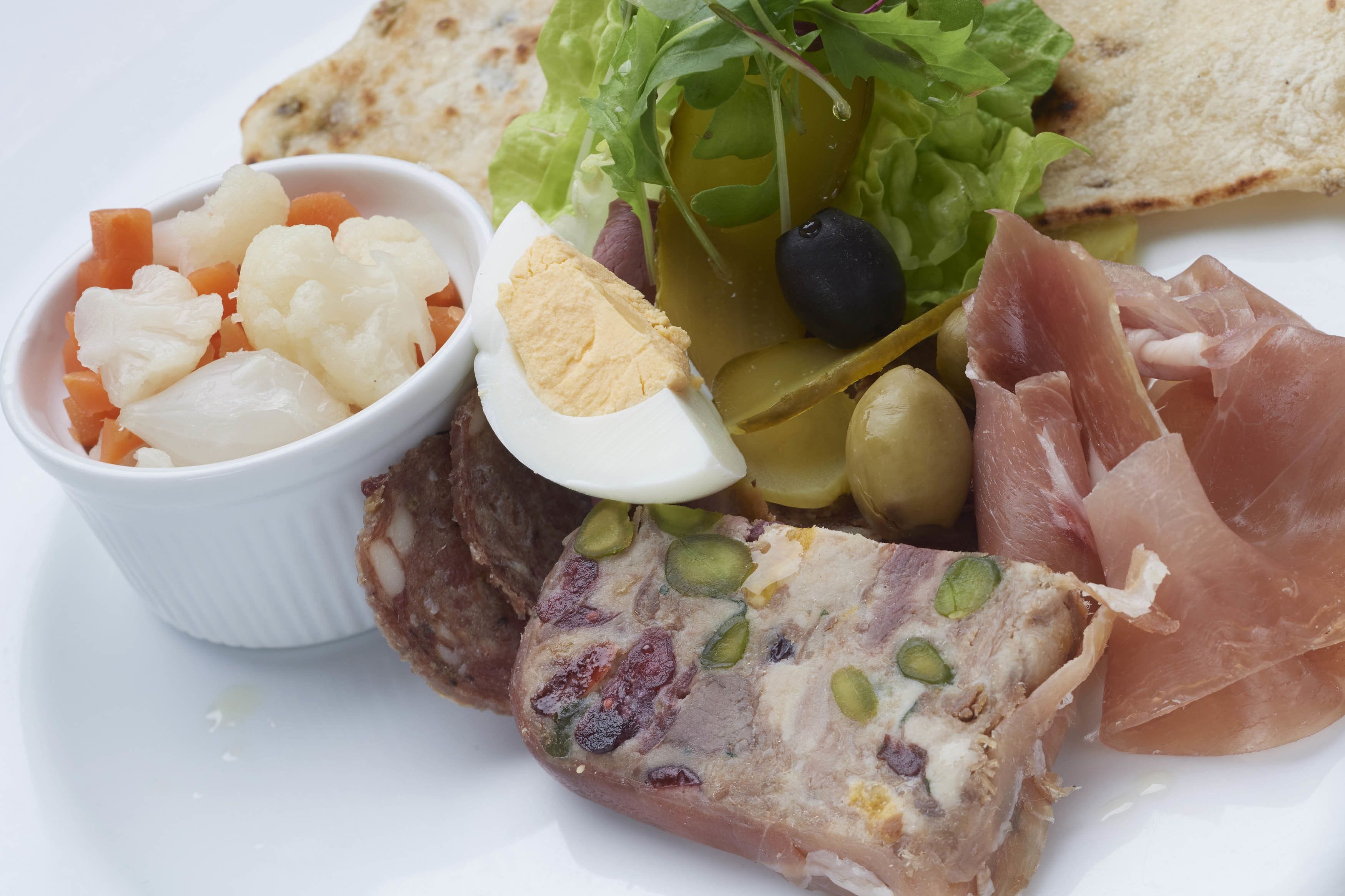 Um prato com uma variedade de alimentos, incluindo uma tigela de legumes picados, uma fatia de carne, azeitonas e um ovo cozido.