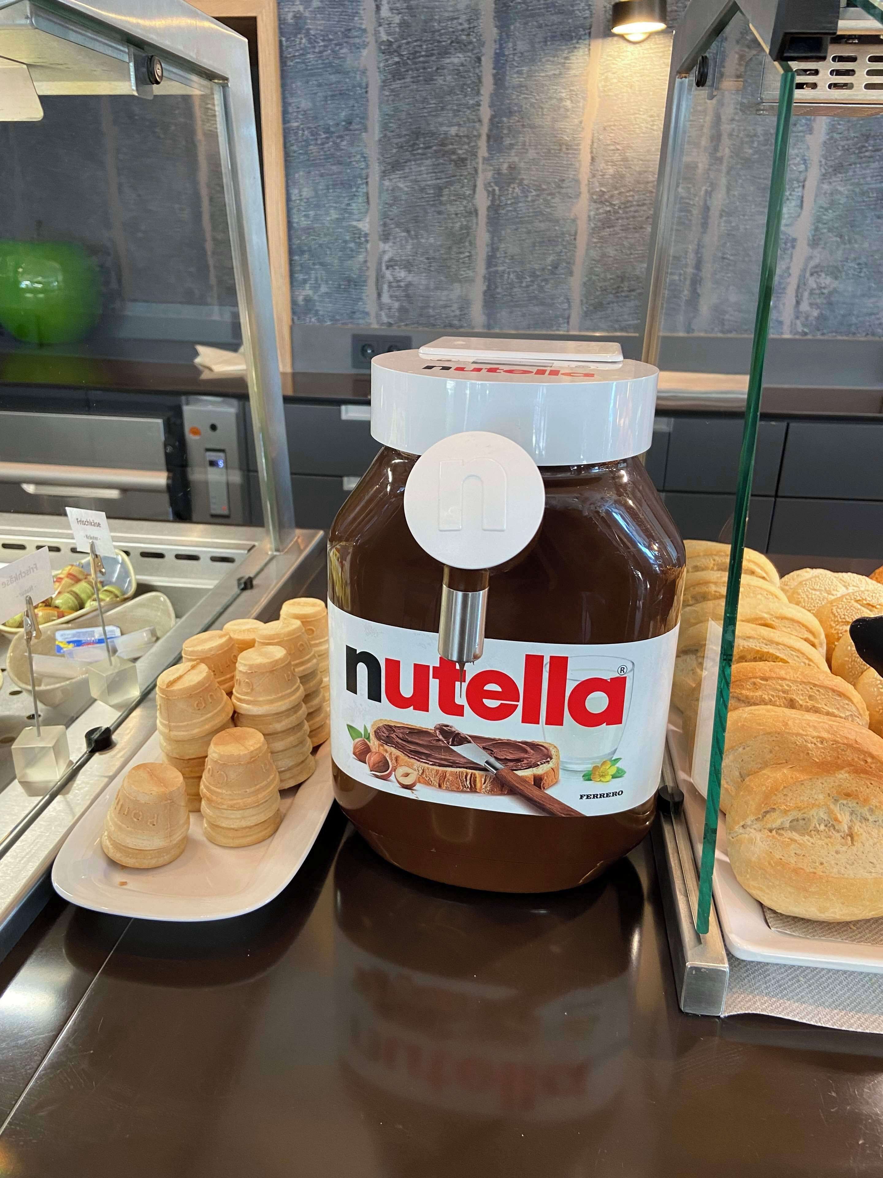 Um pote de Nutella com tampa branca e etiqueta, colocado sobre um balcão ao lado de uma exposição de doces e pão.