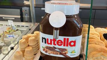 Um pote de Nutella com tampa branca e etiqueta, colocado sobre um balcão ao lado de uma exposição de doces e pão.