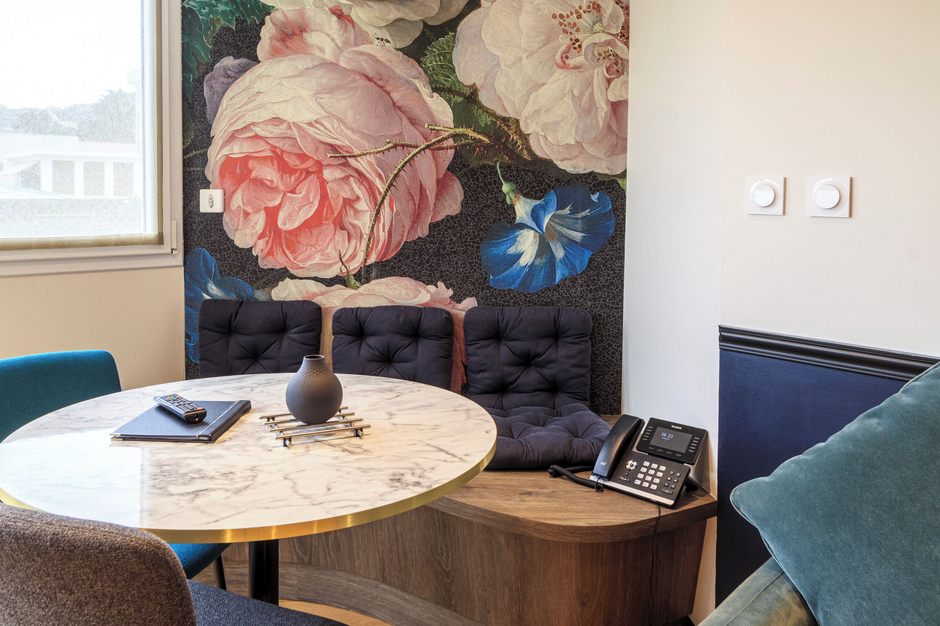 Quarto com mesa de mármore redonda, sofá estofado, pintura floral grande, telefone e janela com vista.