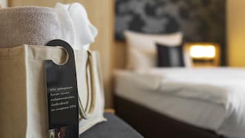 Cama bem arrumada com lençóis brancos, travesseiro cinza e bolsa bege com etiqueta.