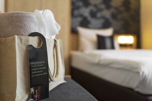 Cama bem arrumada com lençóis brancos, travesseiro cinza e bolsa bege com etiqueta.