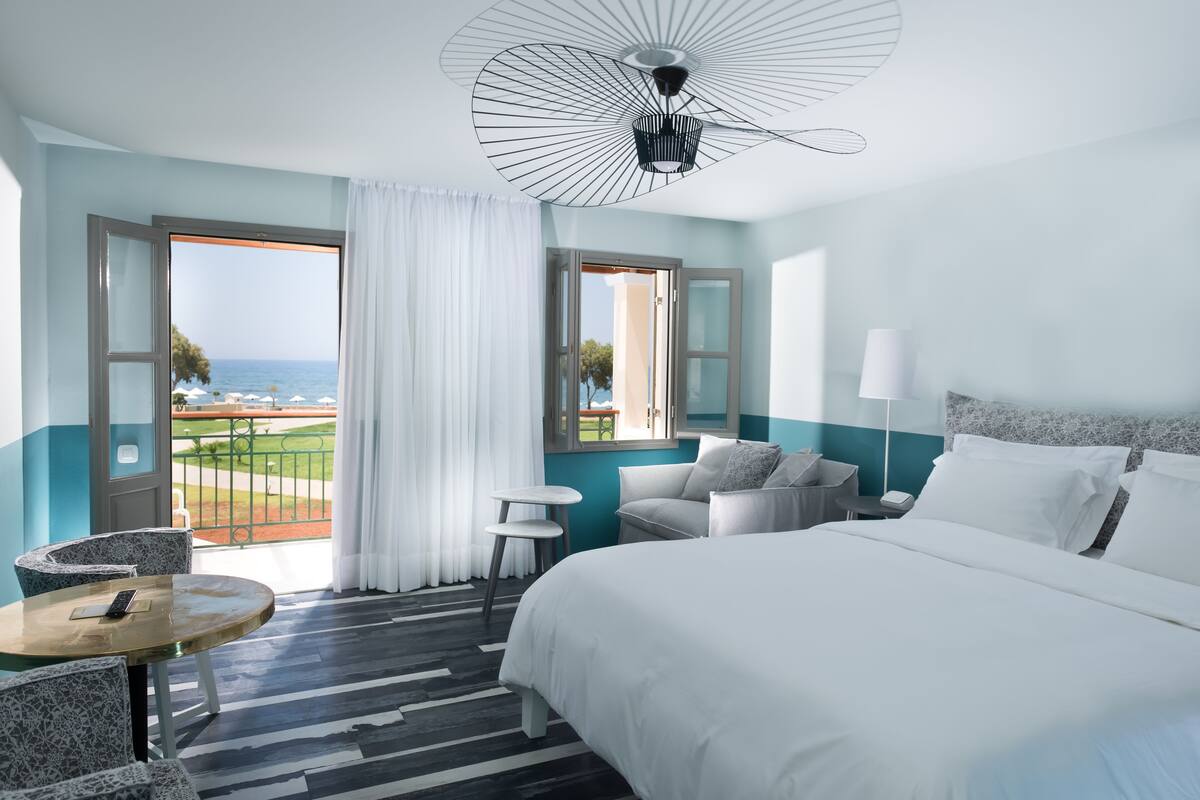 Quarto de hotel com cama, uma pequena mesa, uma cadeira, ventilador de teto e vista para o mar.