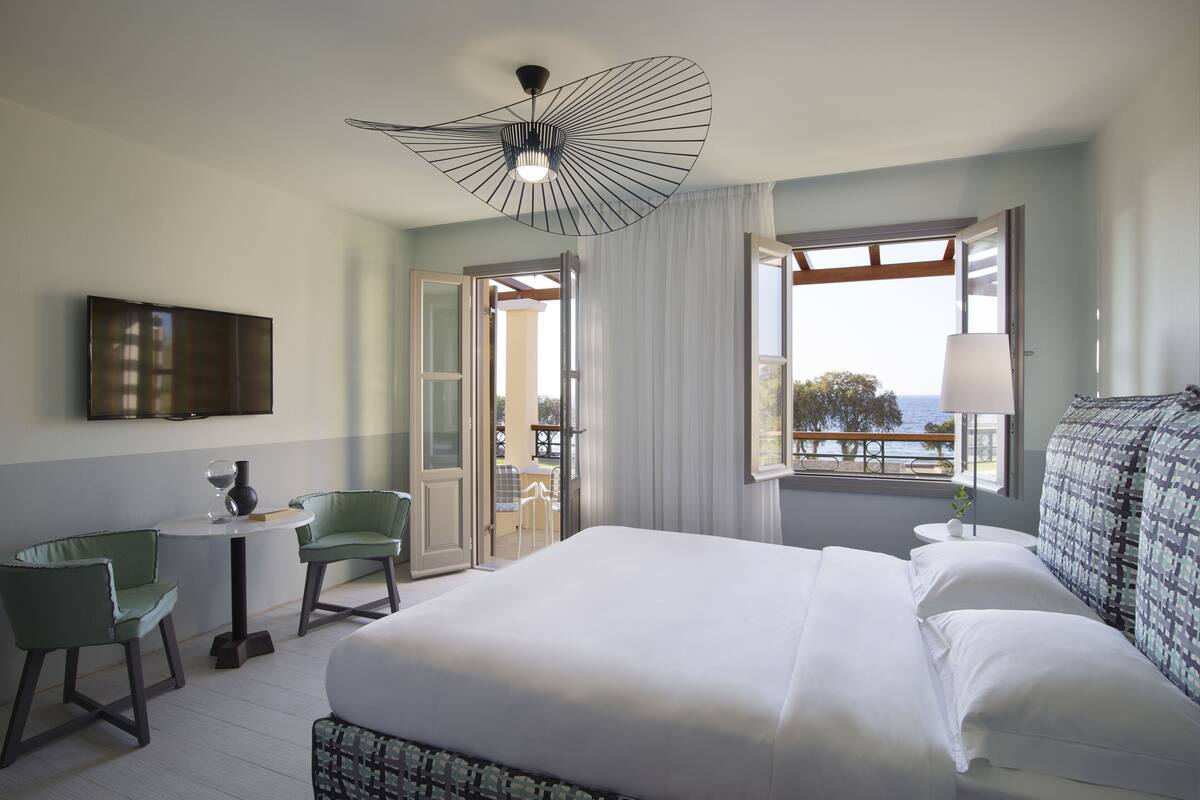 Quarto de hotel com cama grande, televisão, mesa pequena, duas cadeiras e varanda com vista para o mar.
