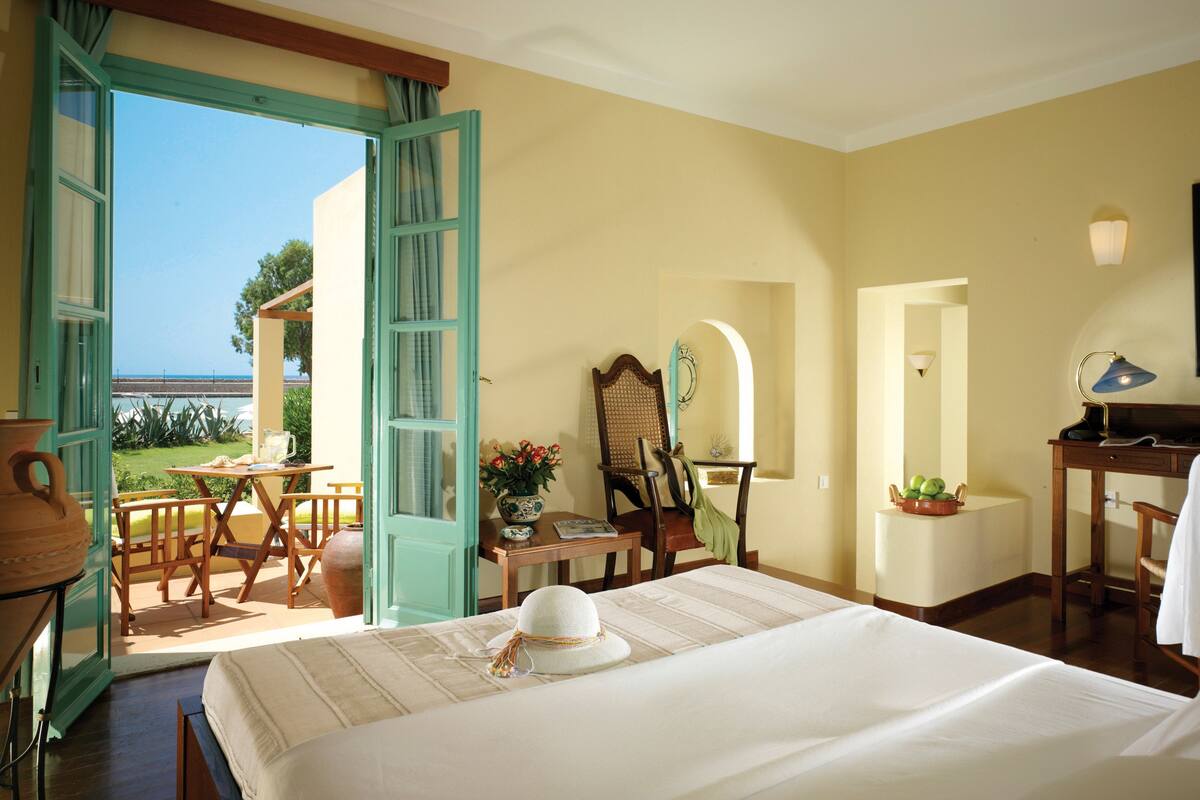 Quarto de hotel com uma cama, uma escrivaninha, uma cadeira, uma mesa e uma porta aberta para uma varanda com vista para o mar.