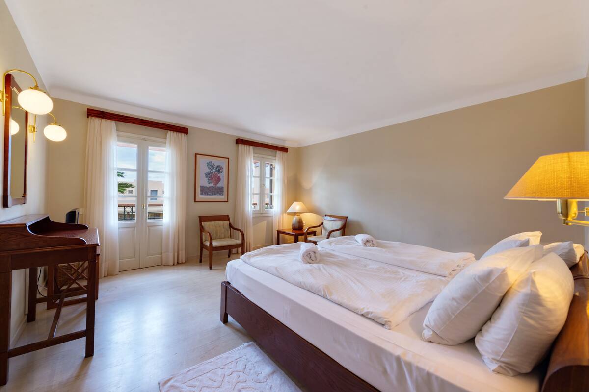 Quarto de hotel com uma cama grande, uma escrivaninha de madeira, duas cadeiras, uma janela com cortinas e um quadro na parede.